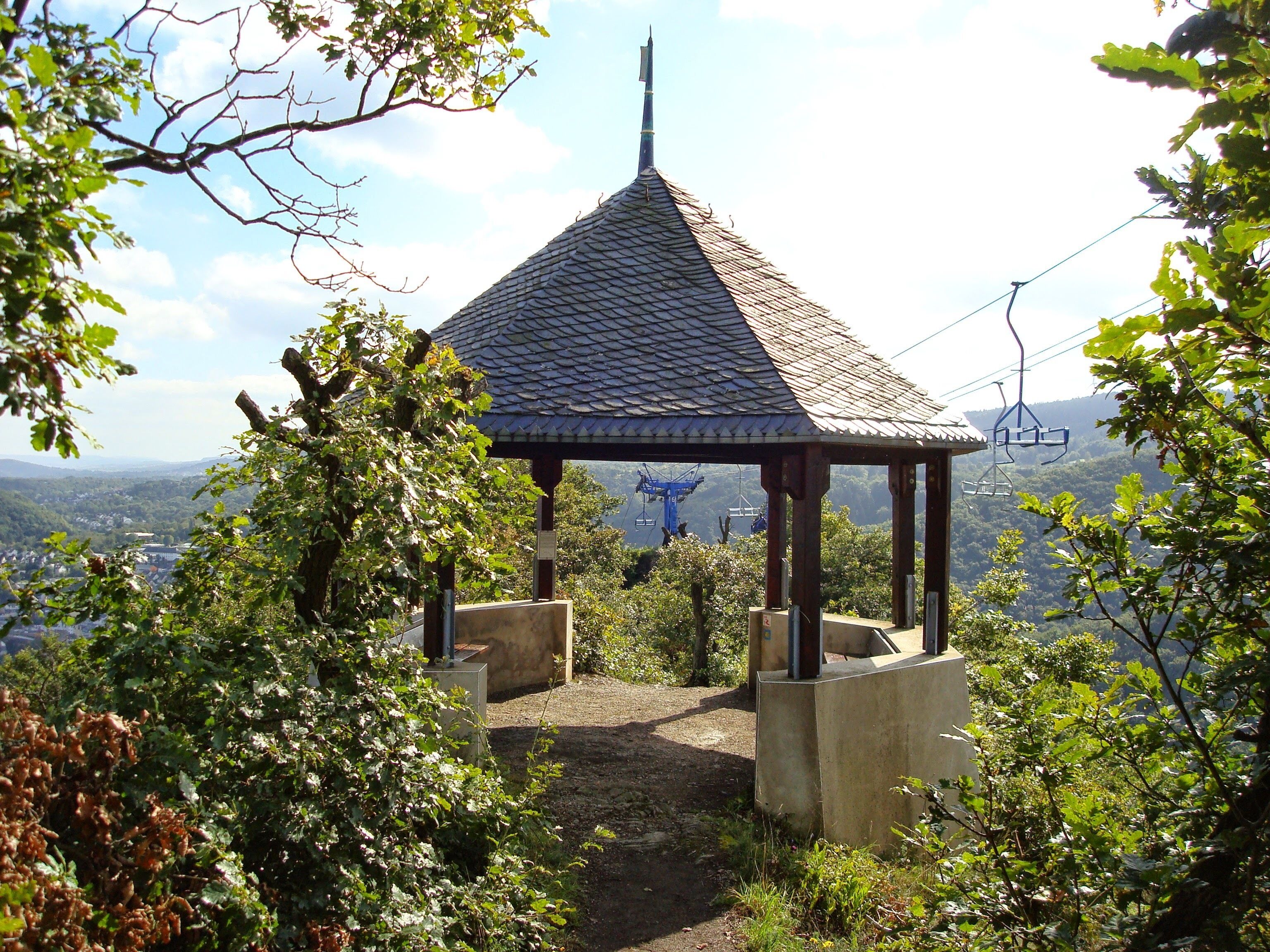 Um gazebo com telhado de azulejos, rodeado por árvores e uma vista distante de uma cordilheira.