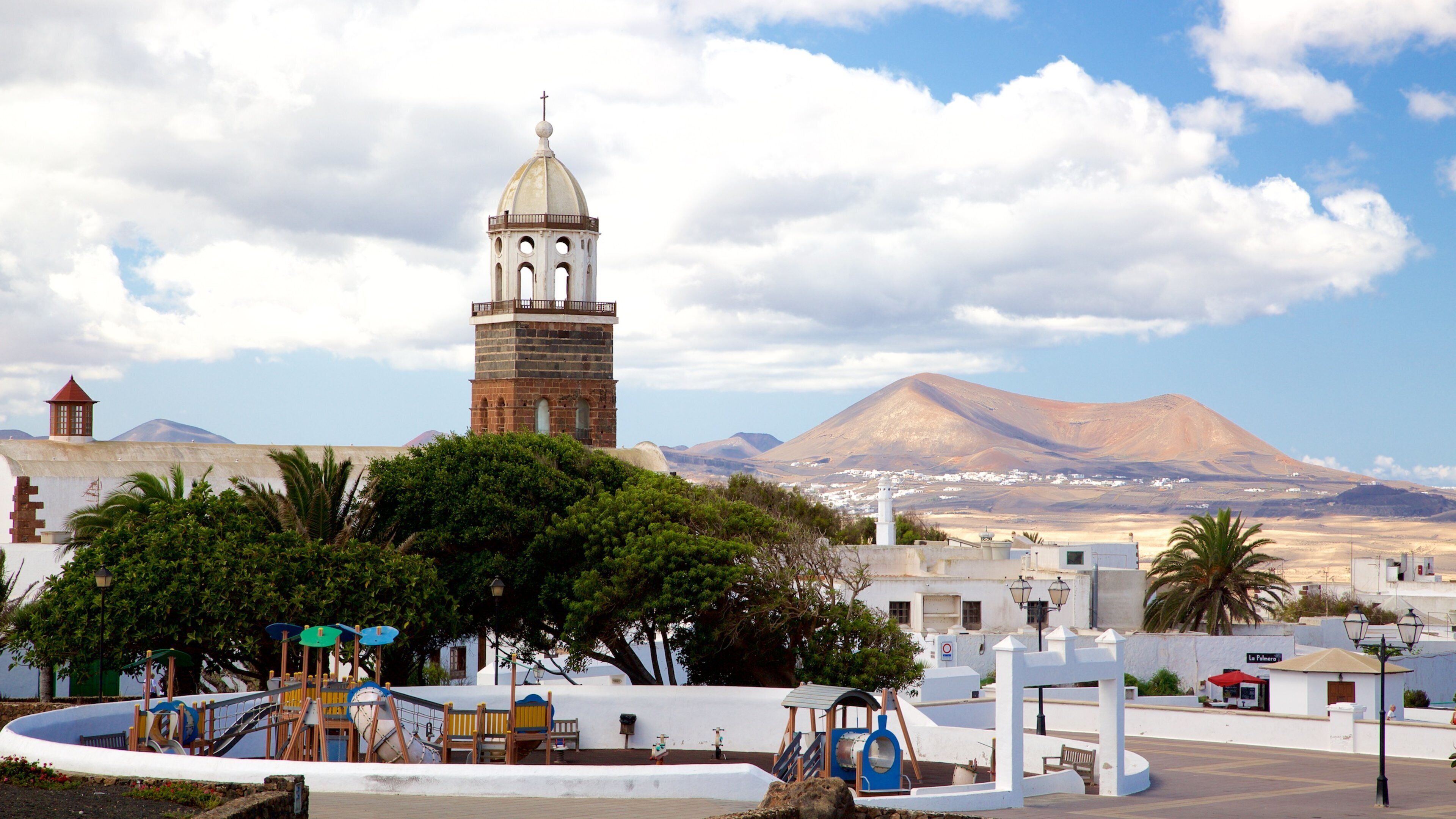 Um parquinho com baloiços e escorregas, um edifício branco com uma torre de sino, e uma montanha ao fundo.
