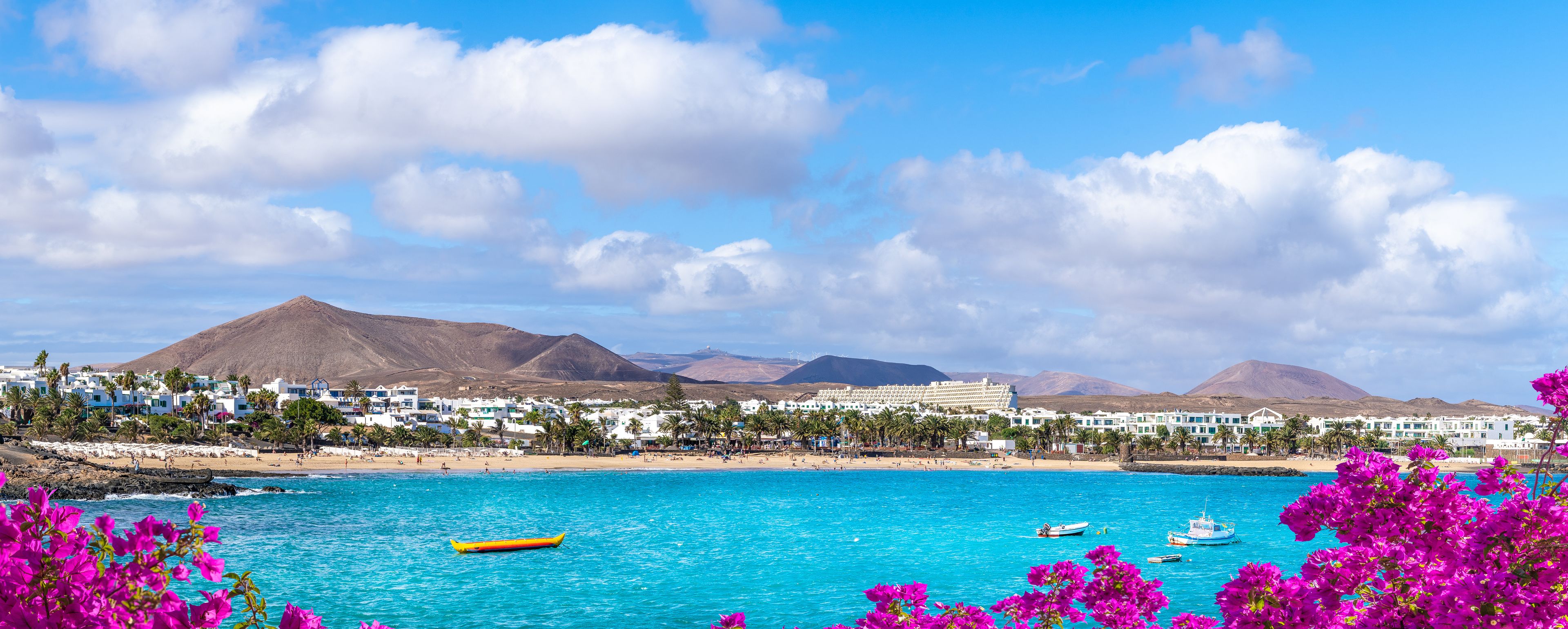 Uma cidade costeira com mar azul cristalino, barcos e montanhas ao fundo.