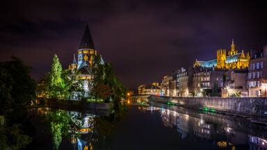Vista noturna de um castelo com uma torre, refletido em um rio calmo, rodeado por edifícios iluminados.