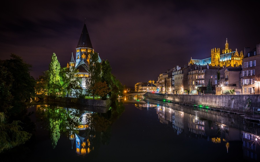 Vista noturna de um castelo com uma torre, refletido em um rio calmo, rodeado por edifícios iluminados.