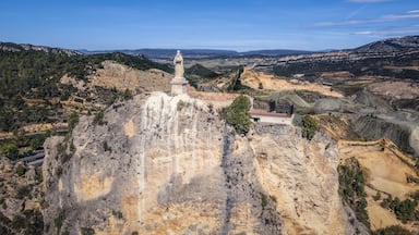 Montanha rochosa com uma estátua no topo, rodeada por um vale e colinas ao longe.