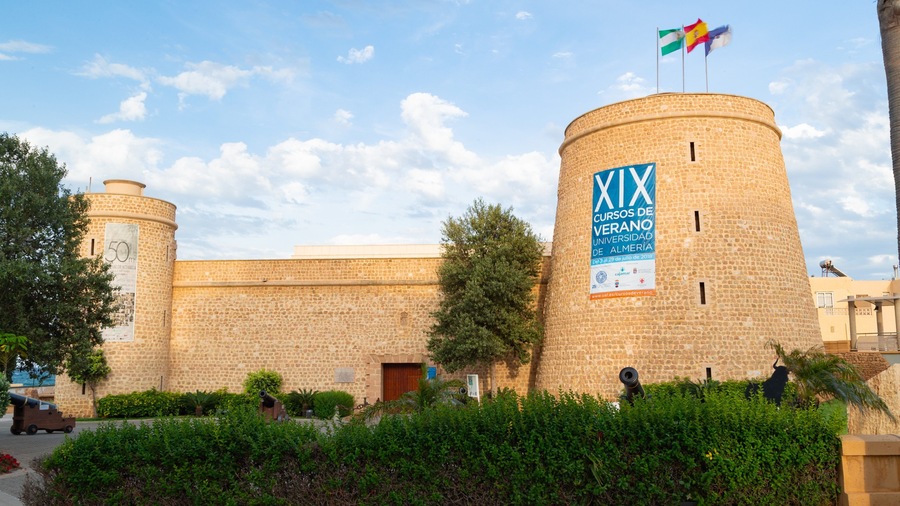 Um edifício histórico de pedra com uma bandeira, um canhão e vegetação exuberante.