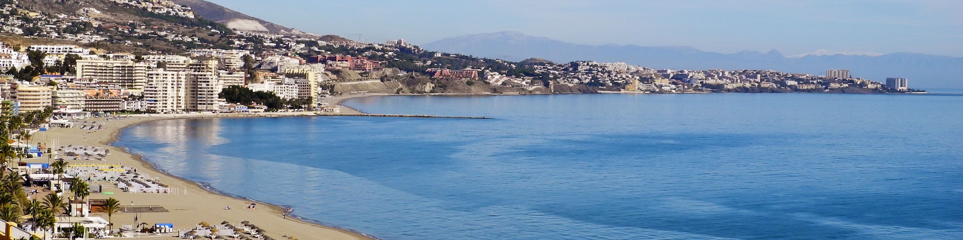 Uma cidade costeira com praia, edifícios e montanhas ao fundo.