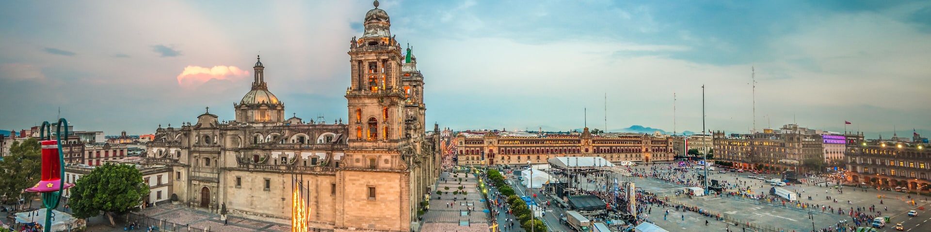 Uma catedral histórica com uma torre de sino imponente, uma praça ampla e aberta com pessoas e veículos, e edifícios urbanos ao redor.