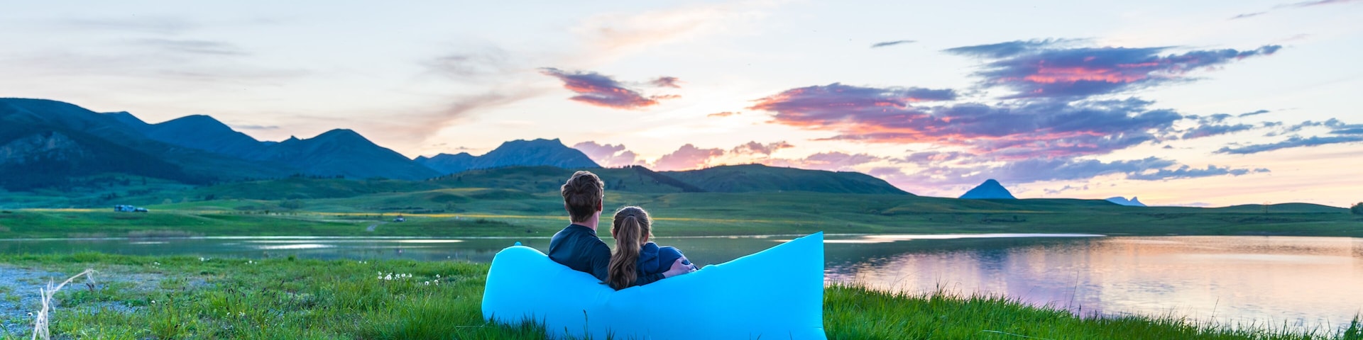 Um casal em um canoa azul em um campo gramado, com um lago e montanhas ao fundo.