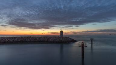 Cena costeira com um farol, um molhe de pedra e águas calmas ao pôr do sol.