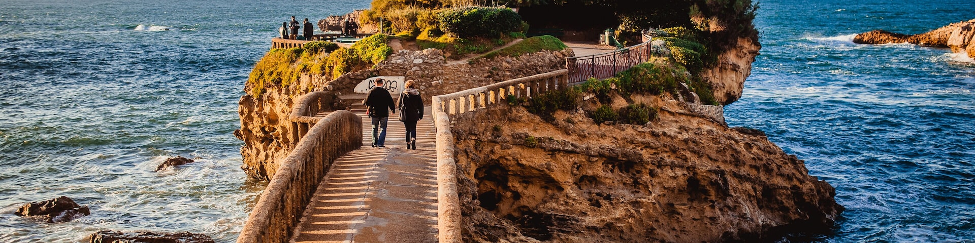 Uma passarela costeira que leva a uma ilha rochosa, com pessoas caminhando.