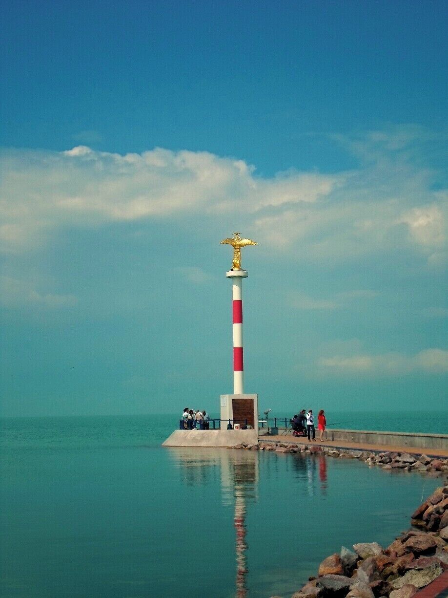 Um farol com uma torre listrada em vermelho e branco e um topo dourado, localizado à beira de um mar tranquilo sob um céu claro.