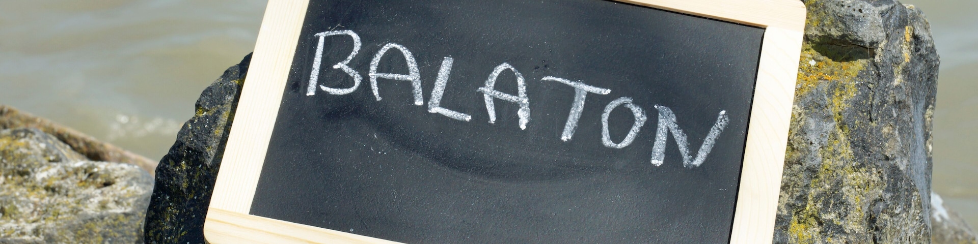 Um quadro negro com a palavra 'BALATON' escrita, apoiado em uma rocha à beira da água.