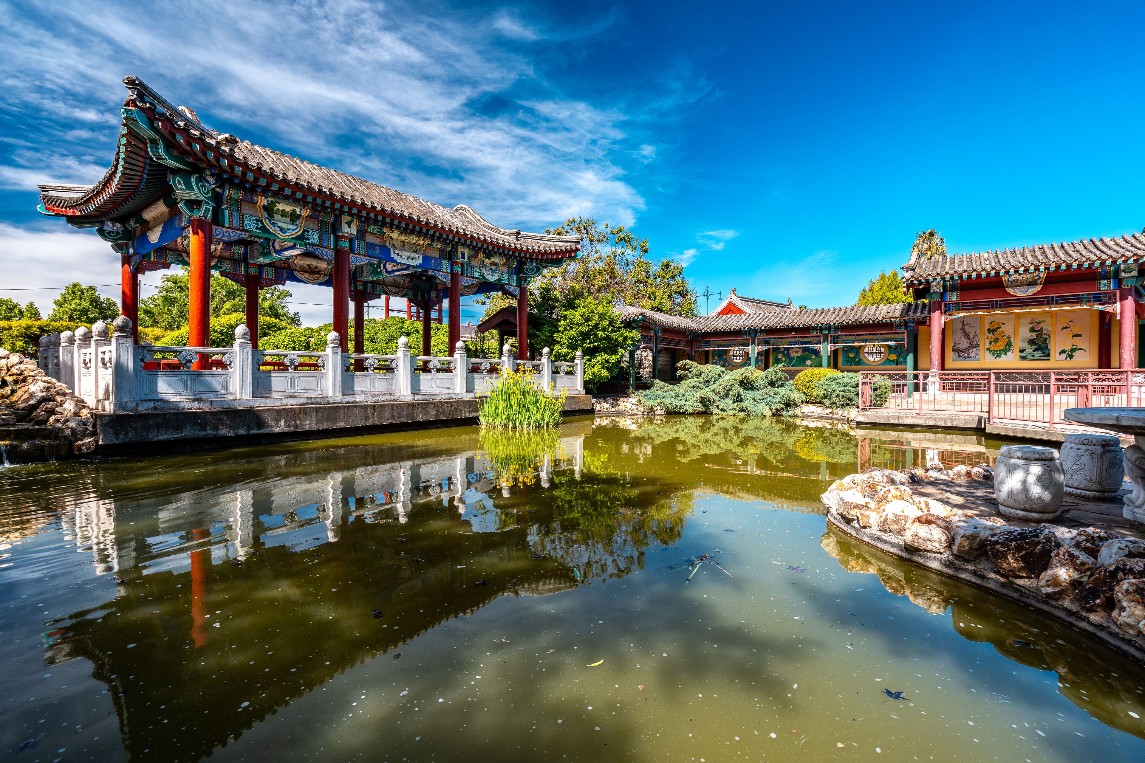 Um pavilhão tradicional chinês sobre um lago com reflexo, rodeado por vegetação exuberante e um céu azul límpido.
