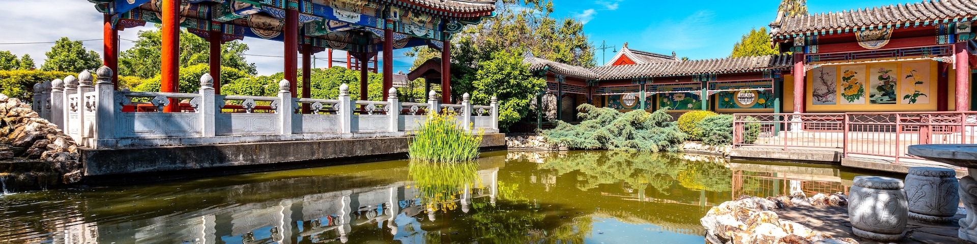 Um pavilhão tradicional chinês sobre um lago com reflexo, rodeado por vegetação exuberante e um céu azul límpido.
