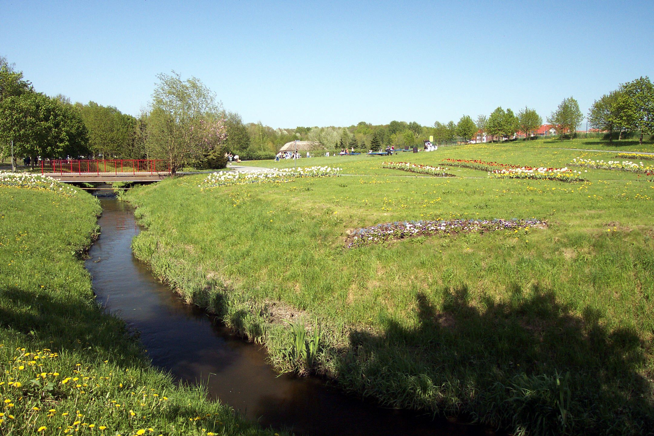 Um rio serpenteando por um parque, com uma ponte, canteiros de flores e árvores.