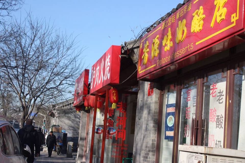 Rua com arquitetura tradicional chinesa, incluindo uma loja com letreiros vermelhos e uma placa indicando uma loja de sobremesas.