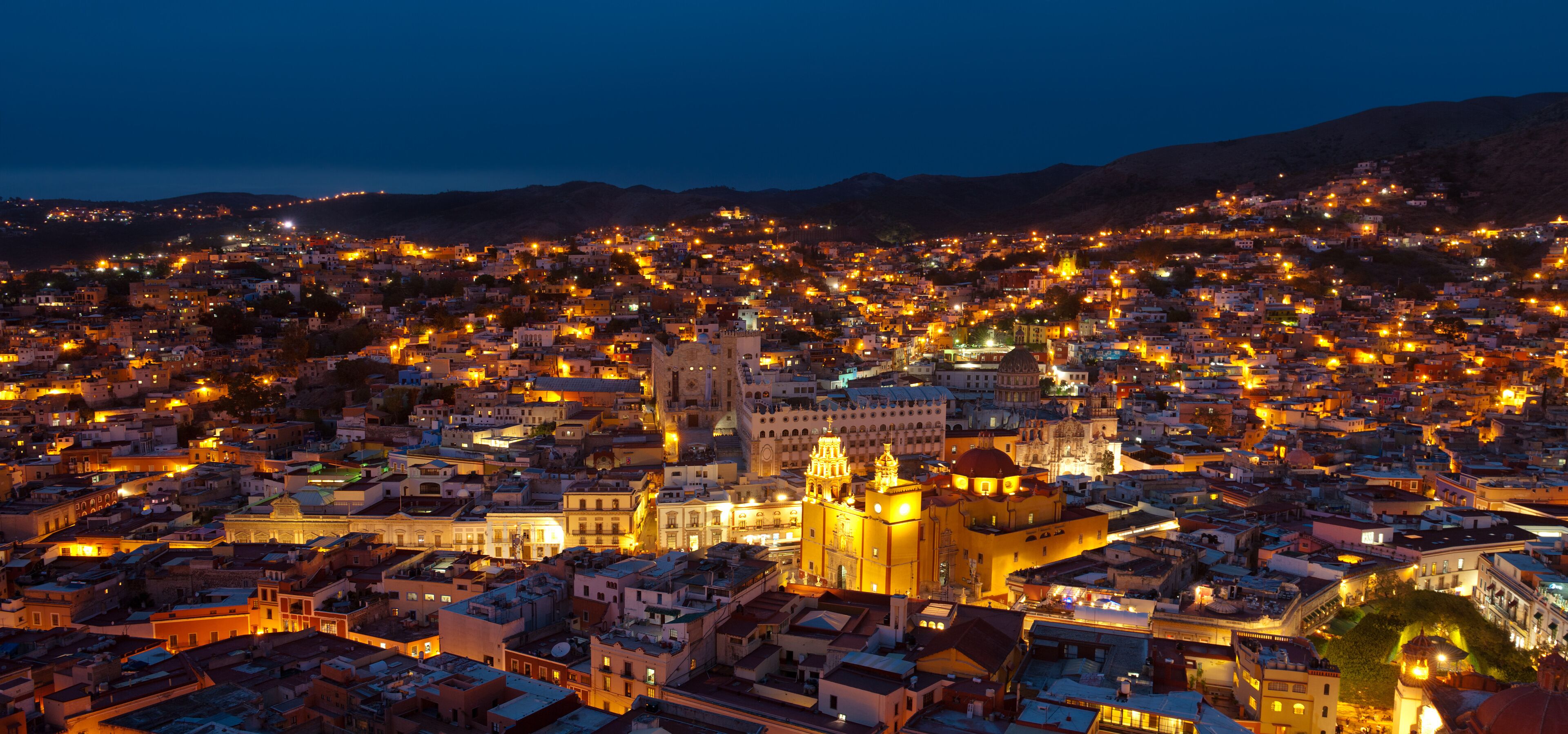 Paisagem urbana ao entardecer, com edifícios iluminados e uma catedral imponente com cúpula.