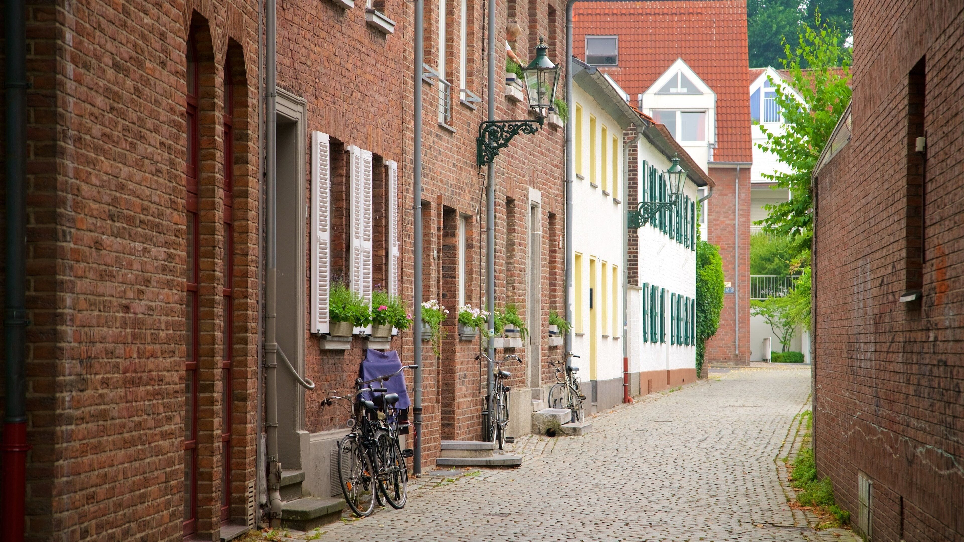 Rua de paralelepípedos ladeada por edifícios de tijolo, alguns com persianas e caixas de flores.