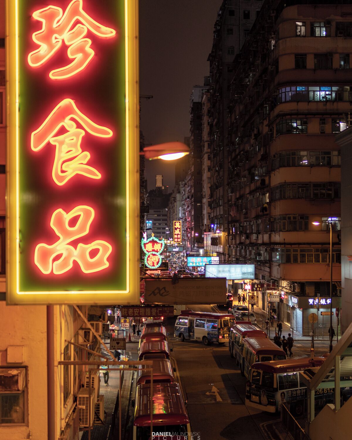 Um letreiro de néon com caracteres chineses, uma rua movimentada com ônibus e edifícios altos à noite.