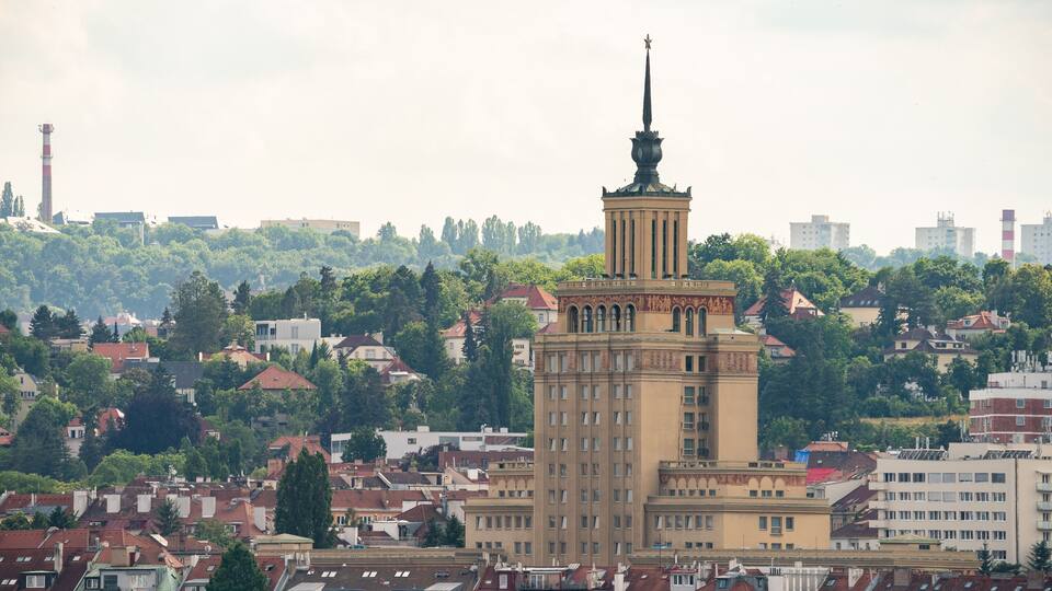 Paisagem urbana com uma torre imponente, tendo uma agulha e uma estátua no topo, cercada por edifícios residenciais e áreas verdes.