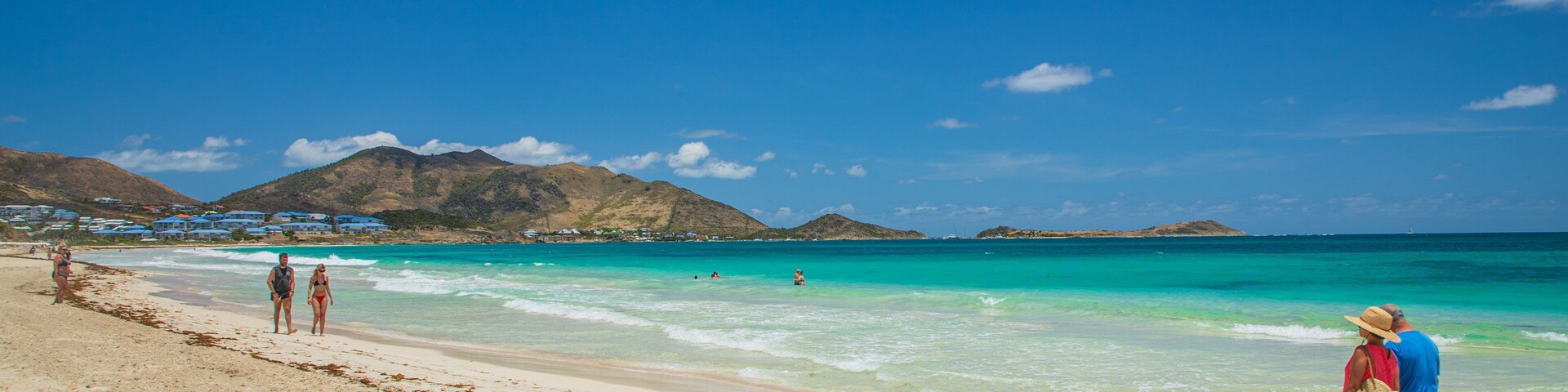 Uma praia com águas turquesa cristalinas, areal arenoso e montanhas ao longe.