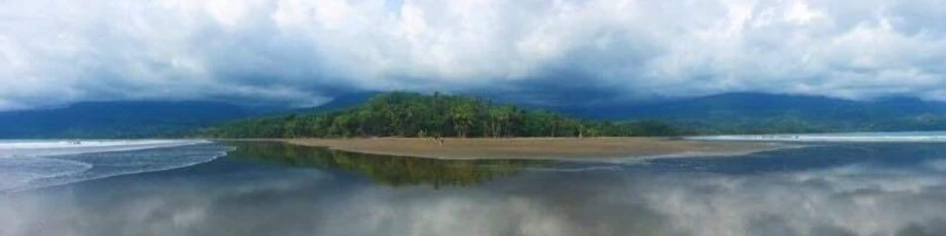 Uma praia com águas calmas refletindo o céu e as nuvens, uma faixa de areia e uma colina arborizada ao longe.