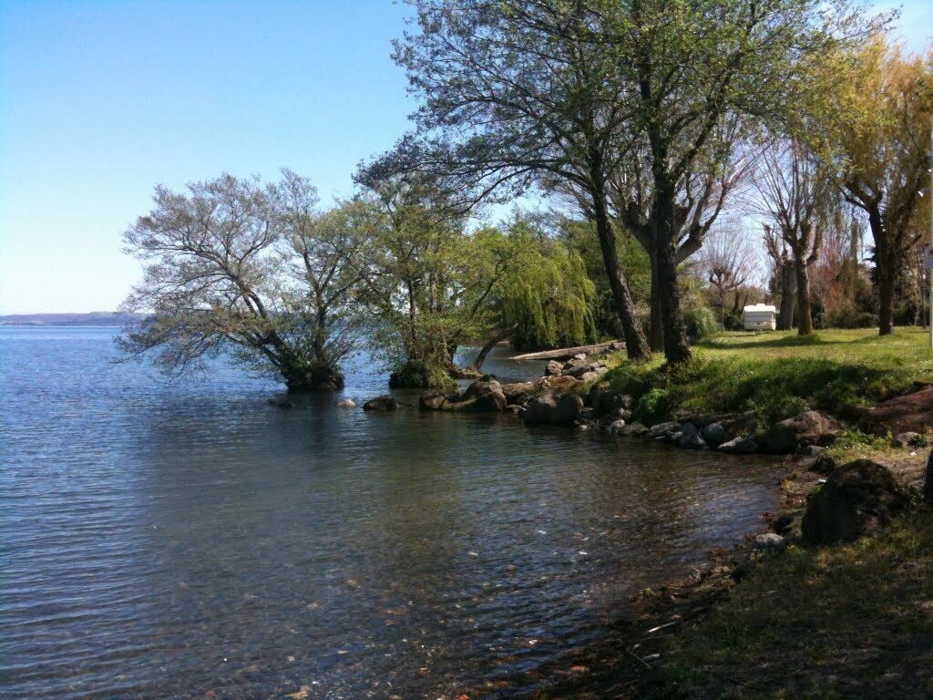 Um lago tranquilo com águas cristalinas, rodeado por árvores e uma área gramada com um pequeno edifício branco.