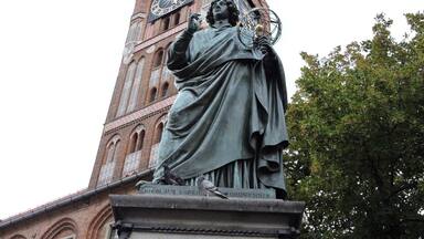 Uma estátua de Nicolau Copérnico segurando um modelo do sistema solar, com uma torre do relógio ao fundo.