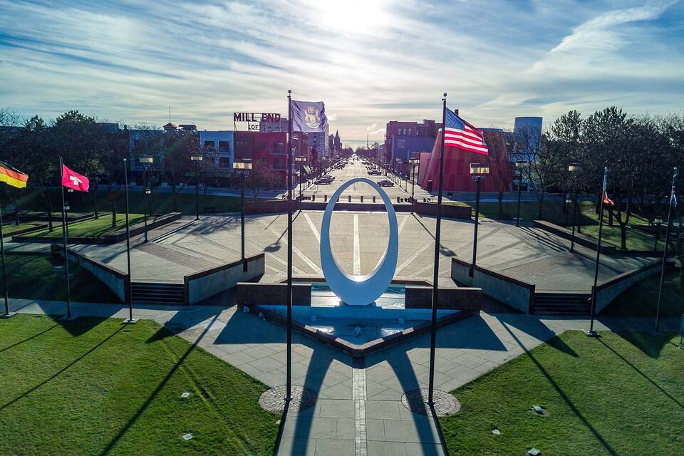 Uma praça com uma grande escultura circular, bandeiras e um edifício com a placa 'MILL END'.