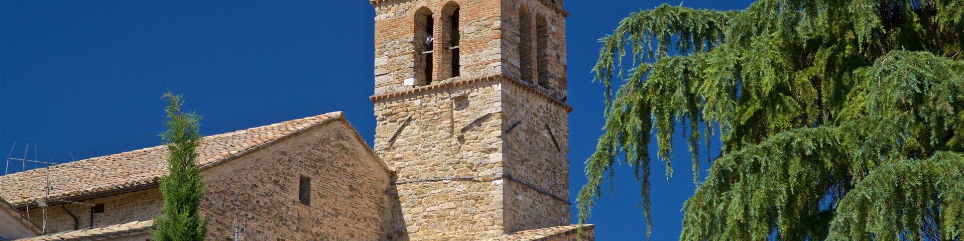 Uma igreja de pedra com uma torre de sino, telhado de telhas e um salgueiro chorão.