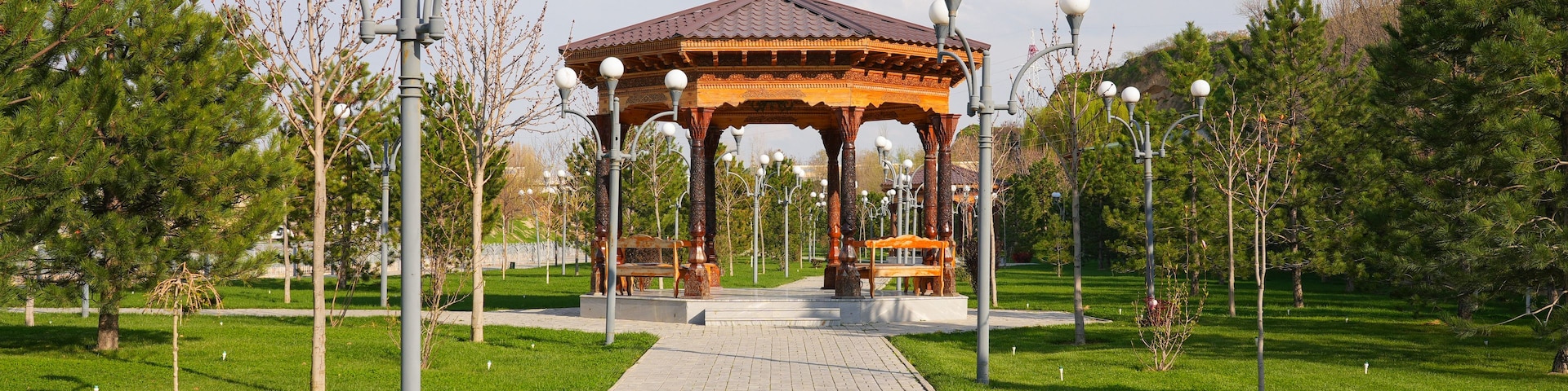 Um gazebo com telhado em telhas e pilares de madeira elaborados, cercado por um caminho pavimentado e postes de luz.