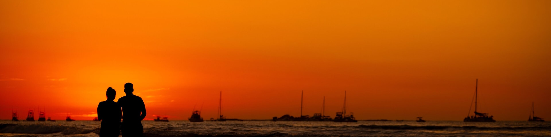 Silhuetas de duas pessoas em uma praia durante o pôr do sol, com barcos na água e um céu laranja vibrante.