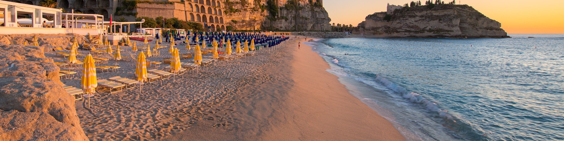 Uma praia costeira com uma fileira de guarda-sóis amarelos, um afloramento rochoso e edifícios históricos ao fundo.