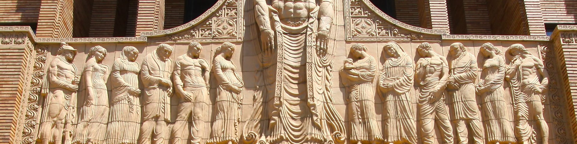 Escultura em baixo-relevo de pedra detalhada em fachada de edifício, com uma figura central acompanhada por figuras menores em diferentes poses.