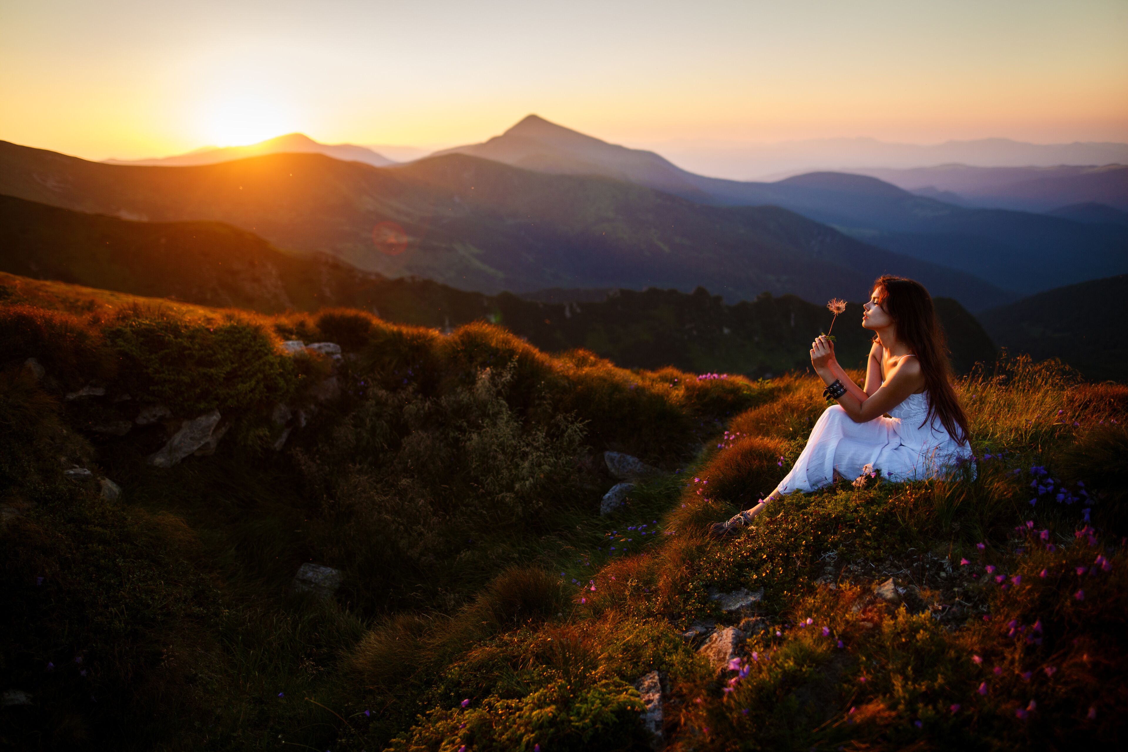 Uma pessoa sentada numa colina gramada com montanhas ao fundo durante o pôr do sol.