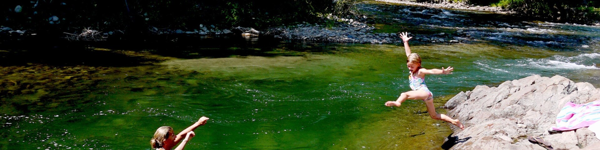 Duas pessoas saltam para um rio límpido, rodeado por árvores.