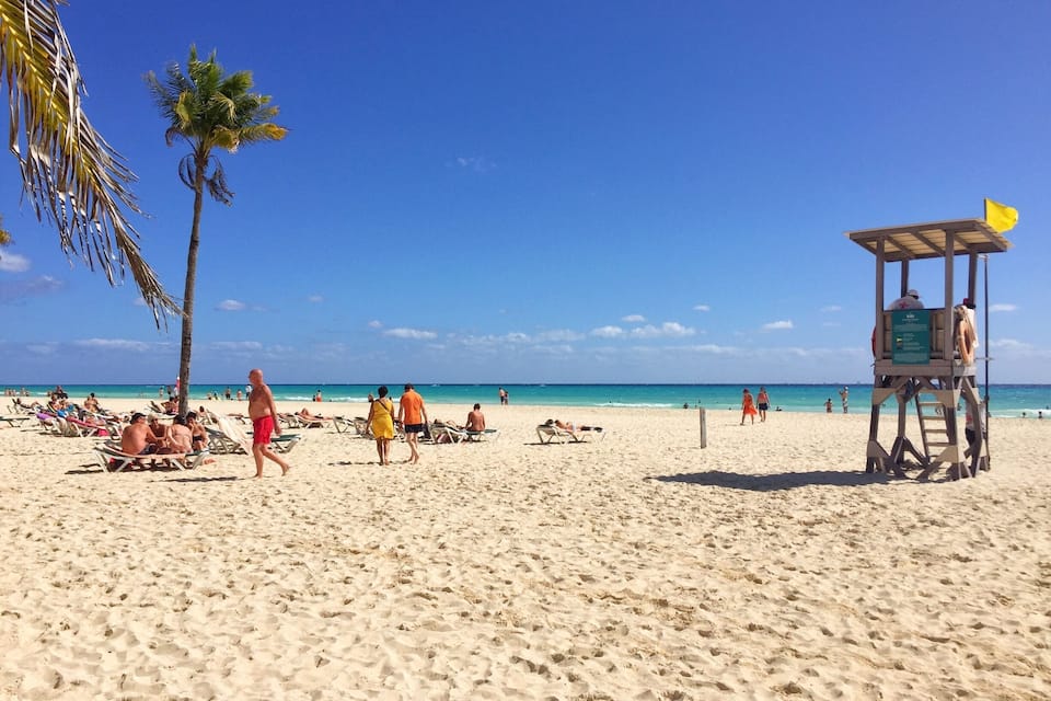 Uma praia arenosa com palmeiras, cadeiras de praia e uma torre de salva-vidas.