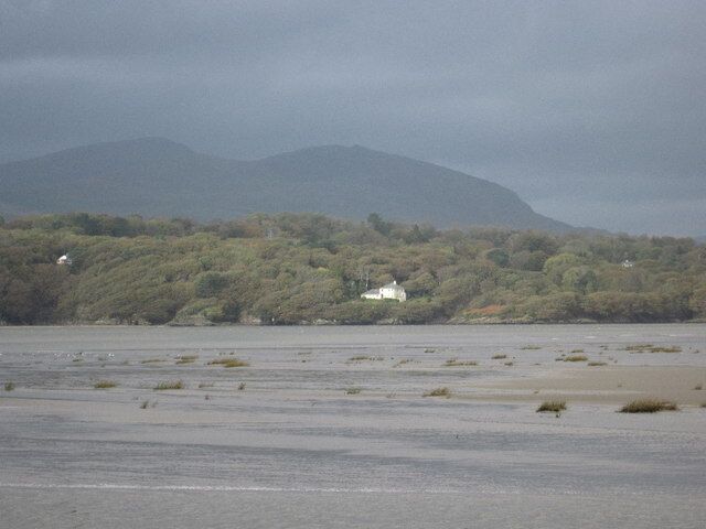 Paisagem costeira com um edifício branco numa colina, rodeado por vegetação densa e montanhas ao fundo.