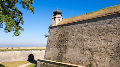 Um muro de pedra com uma torre de vigia, céu claro e um caminho que leva ao muro.