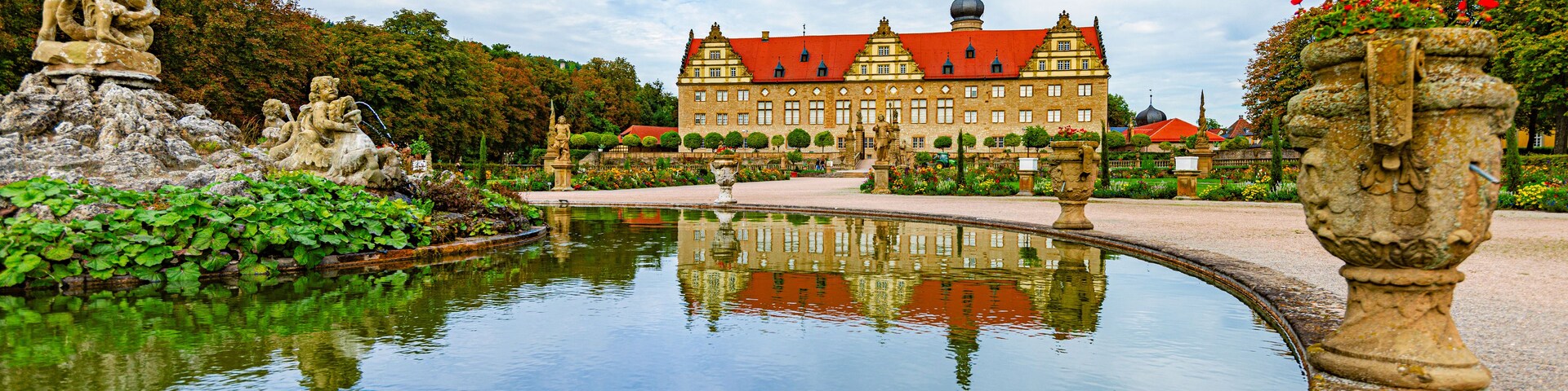 Um edifício histórico com telhado vermelho, um grande lago refletindo a estrutura, e estátuas ao redor do lago.