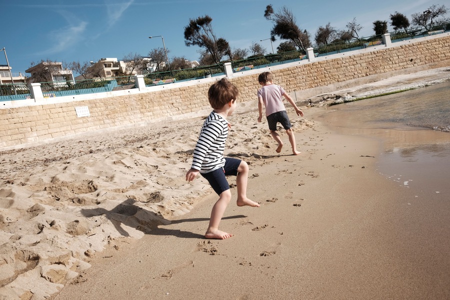 Duas crianças brincando numa praia arenosa, com um muro de pedra e uma cerca ao fundo.