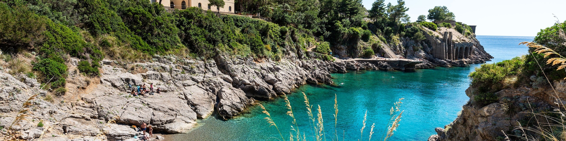 Paisagem costeira com praia, falésias rochosas e um edifício numa encosta.
