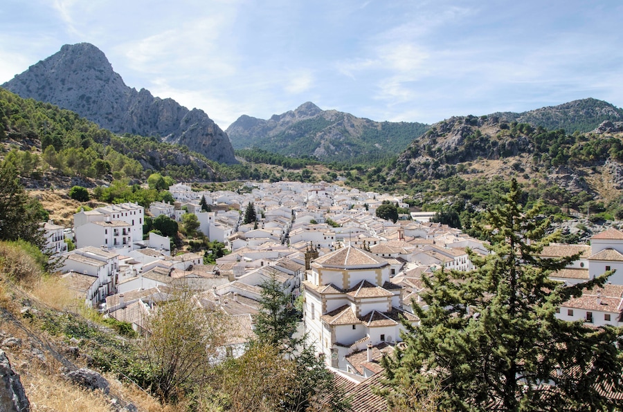 Uma cidade montanhosa com edifícios brancos e telhados de terracota, cercada por colinas verdes exuberantes e um pico rochoso imponente.