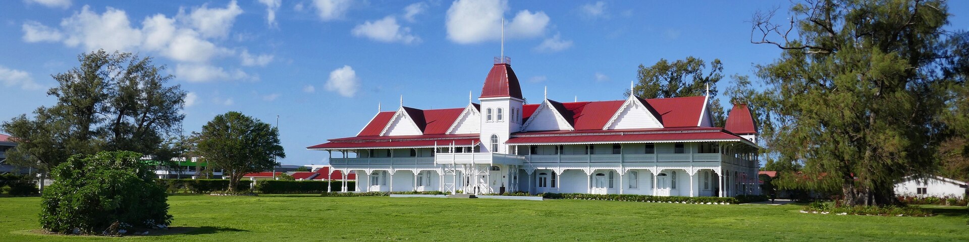 Um edifício grande e branco, com telhado vermelho e várias chaminés, rodeado por um gramado bem cuidado e árvores.