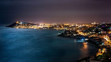 Uma cidade costeira à noite, com uma colina imponente e um litoral curvo.