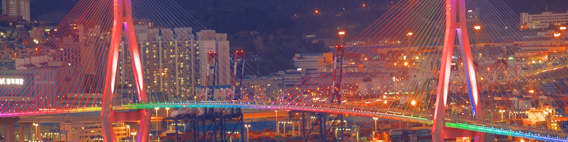 Uma ponte estaiada bem iluminada com luzes coloridas, paisagem urbana ao fundo e um corpo d'água em primeiro plano.