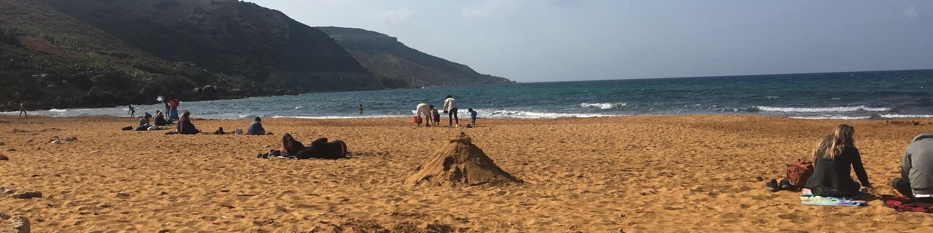 Uma praia de areia com pegadas, algumas pessoas e uma paisagem montanhosa ao fundo.