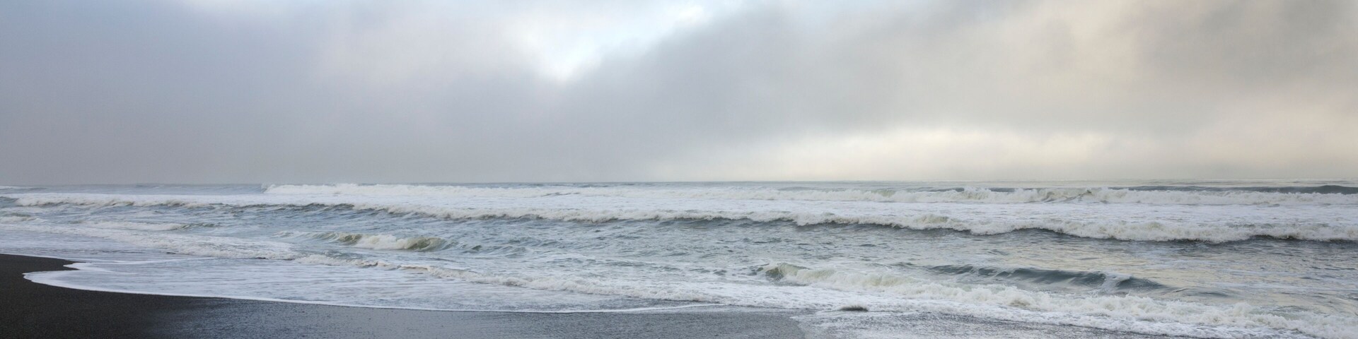 Uma praia com ondas, céu nublado e faixa de areia escura.
