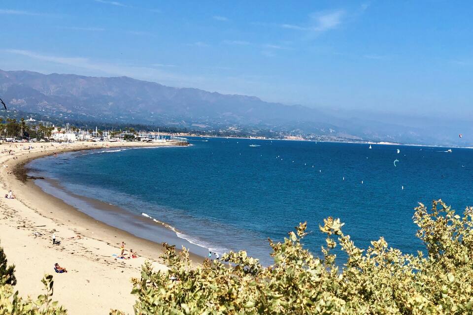 Uma praia costeira com águas azul-claras, areal arenoso e montanhas ao longe.