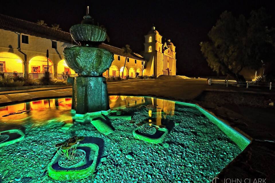 Uma fonte noturna com padrões iluminados, uma igreja com um campanário e um espaço pavimentado com bancos.