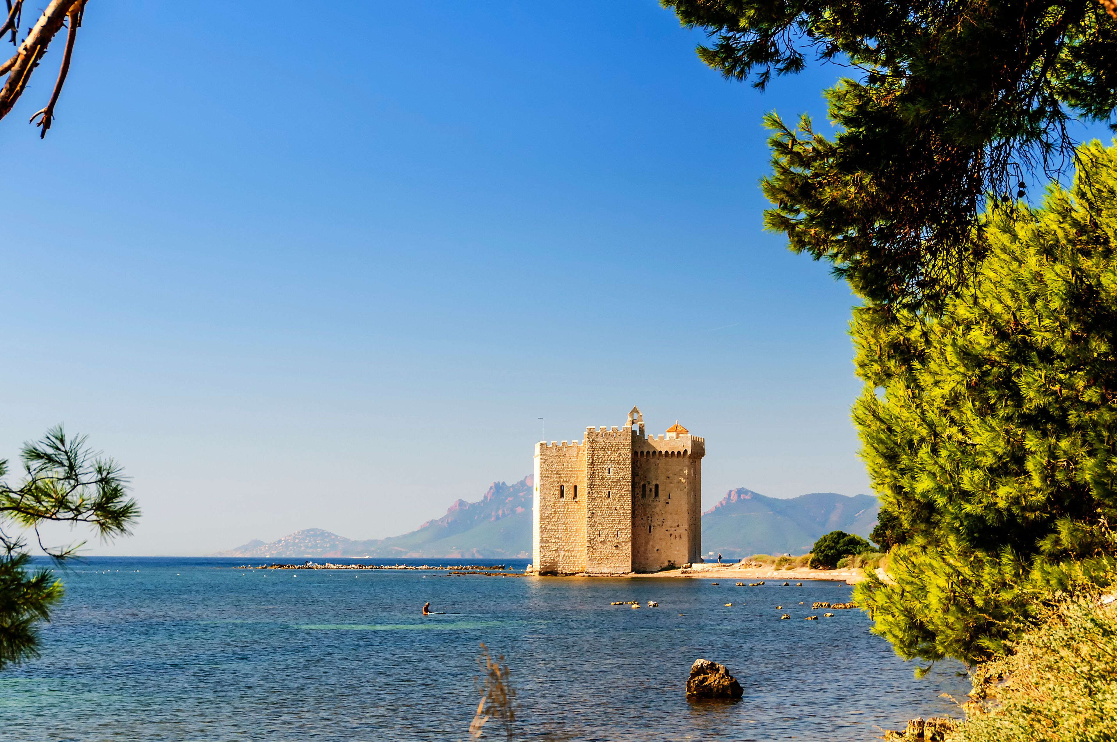 Um castelo à beira-mar, rodeado por águas azuis cristalinas e pinheiros.
