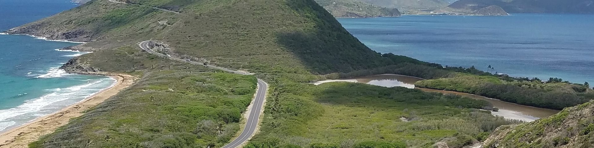 Uma estrada costeira serpenteia por colinas verdes exuberantes, com uma praia e oceano ao fundo.